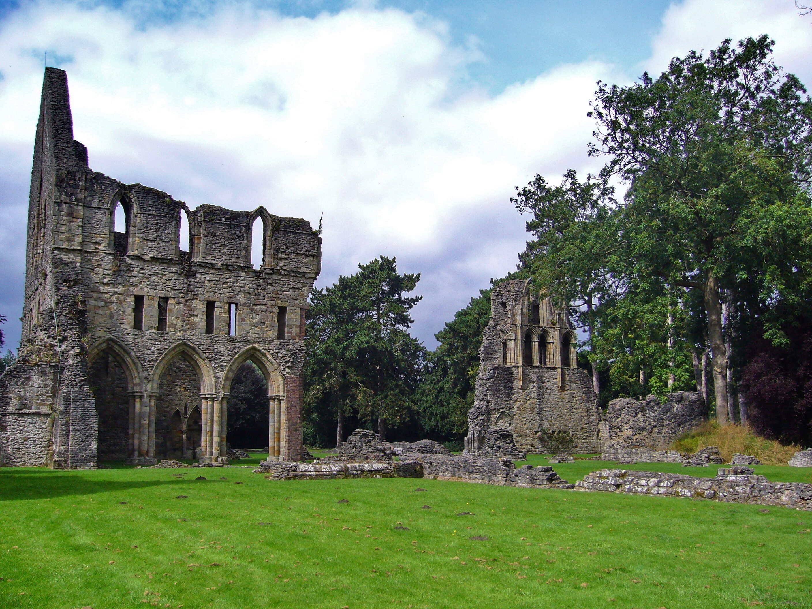 Ruínas de uma estrutura de pedra medieval com arcos e colunas, rodeadas por vegetação exuberante e árvores.