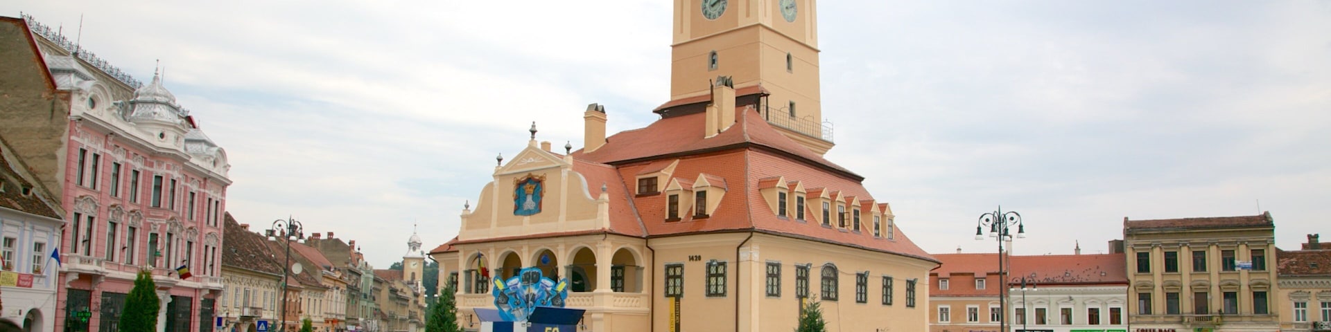 Uma praça histórica com uma torre de igreja imponente, uma fonte central e edifícios ao redor.
