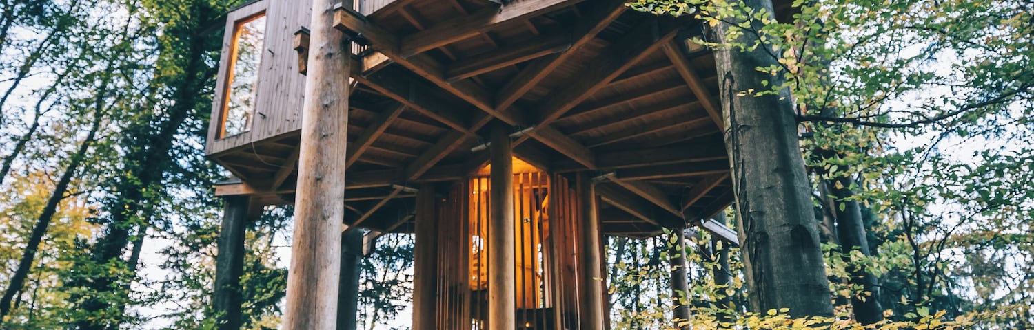 Uma cabana na árvore em meio a uma floresta, com uma mulher em frente.