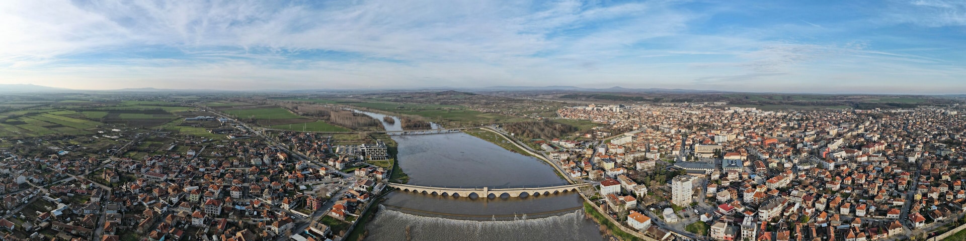 Vista panorâmica de uma cidade com uma ponte imponente sobre um rio, rodeada por campos e colinas ao longe.