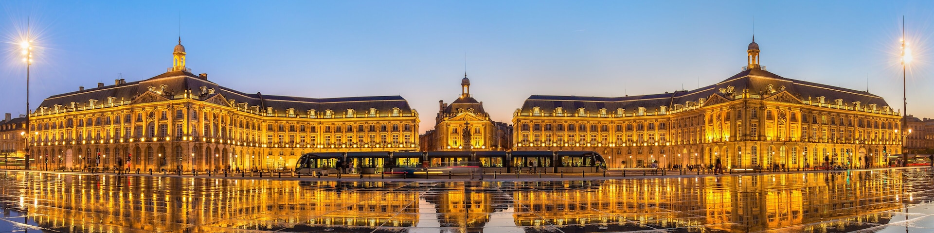 Um edifício imponente e iluminado, com uma cúpula central e duas alas simétricas, refletindo em uma praça molhada.