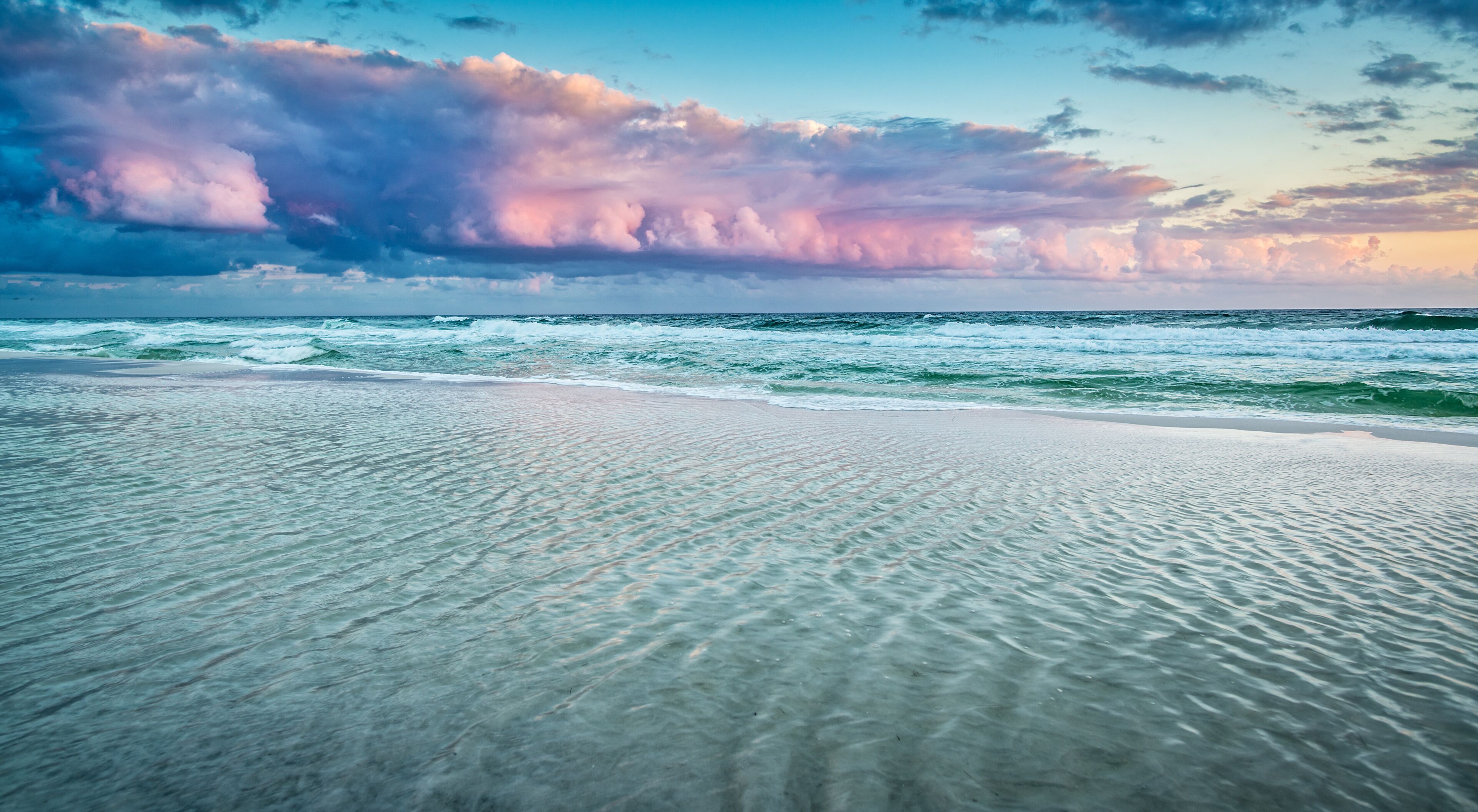 Uma praia com águas cristalinas, areal arenoso e céu dramático com nuvens.