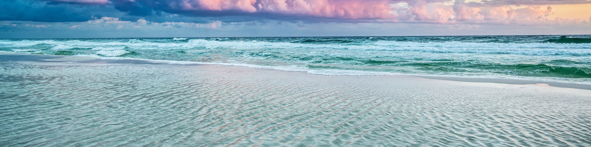 Uma praia com águas cristalinas, areal arenoso e céu dramático com nuvens.