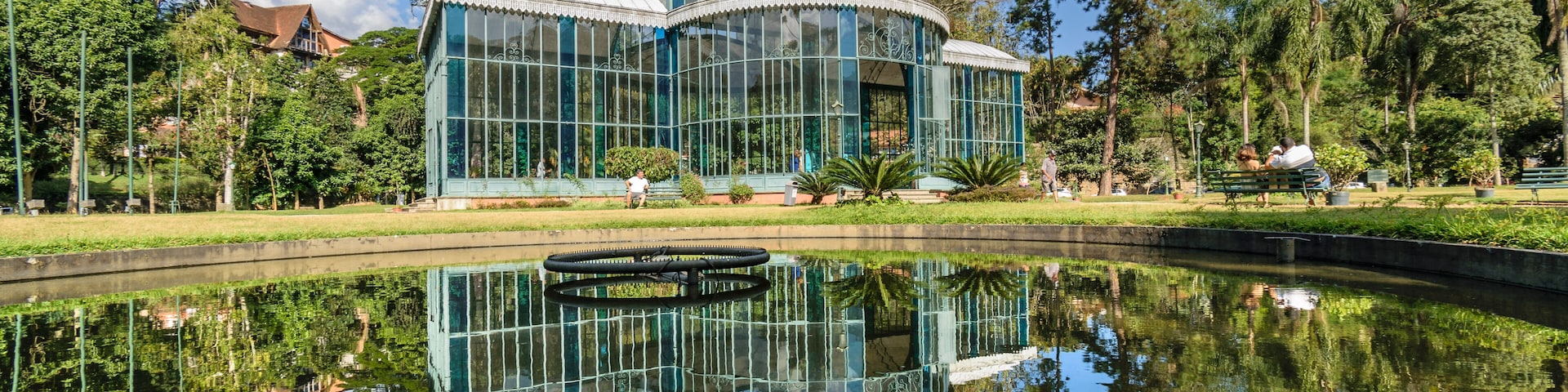 Uma grande estufa de vidro com reflexo em um lago, cercada por árvores e um céu claro.