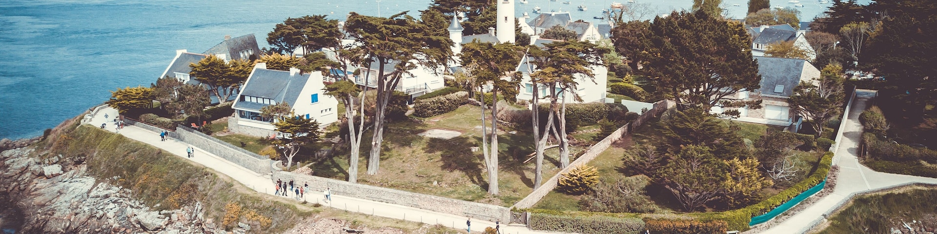 Uma vila costeira com um farol, litoral rochoso e um caminho que leva a uma casa branca com porta azul.