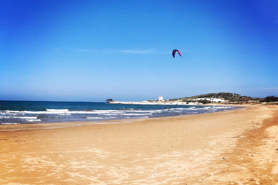 Uma praia com um kitesurfista, ondas e uma colina distante com edifícios.