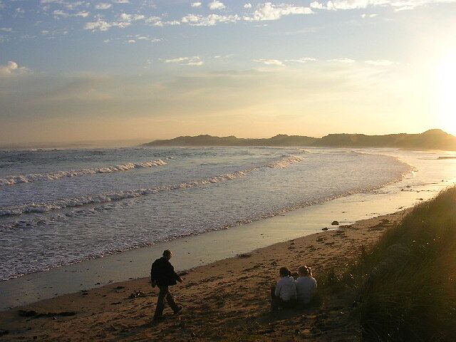 Uma praia com ondas, uma pessoa caminhando e três pessoas sentadas na areia.