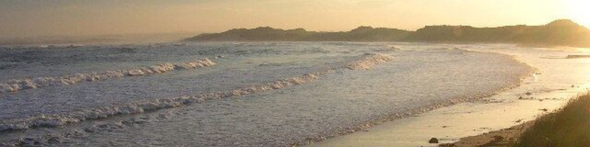 Uma praia com ondas, uma pessoa caminhando e três pessoas sentadas na areia.