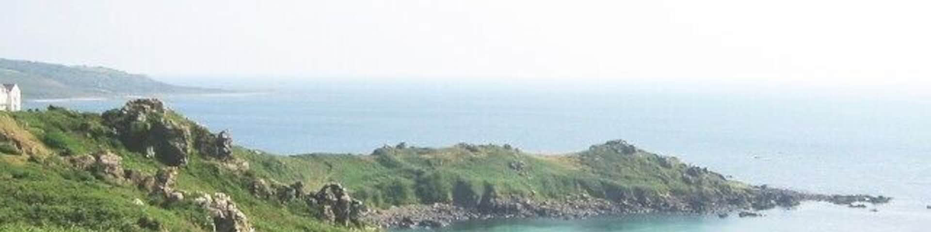 Paisagem costeira com colinas verdes, afloramentos rochosos e mar calmo.