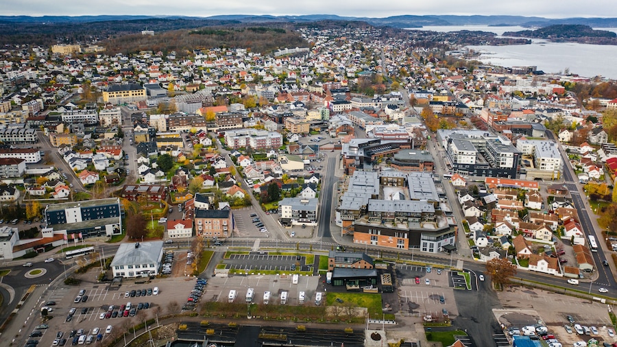 Vista aérea de uma cidade densamente povoada, com uma mistura de edifícios residenciais e comerciais, uma grande massa de água ao fundo e um edifício redondo proeminente no centro.