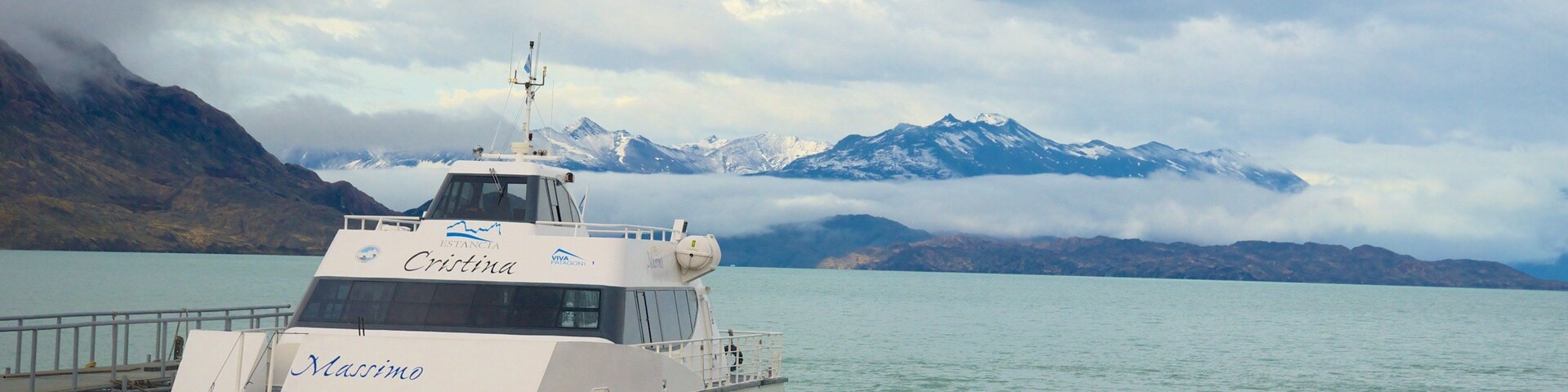 Um barco chamado 'Cristina' atracado em um cais, com uma paisagem montanhosa ao fundo.