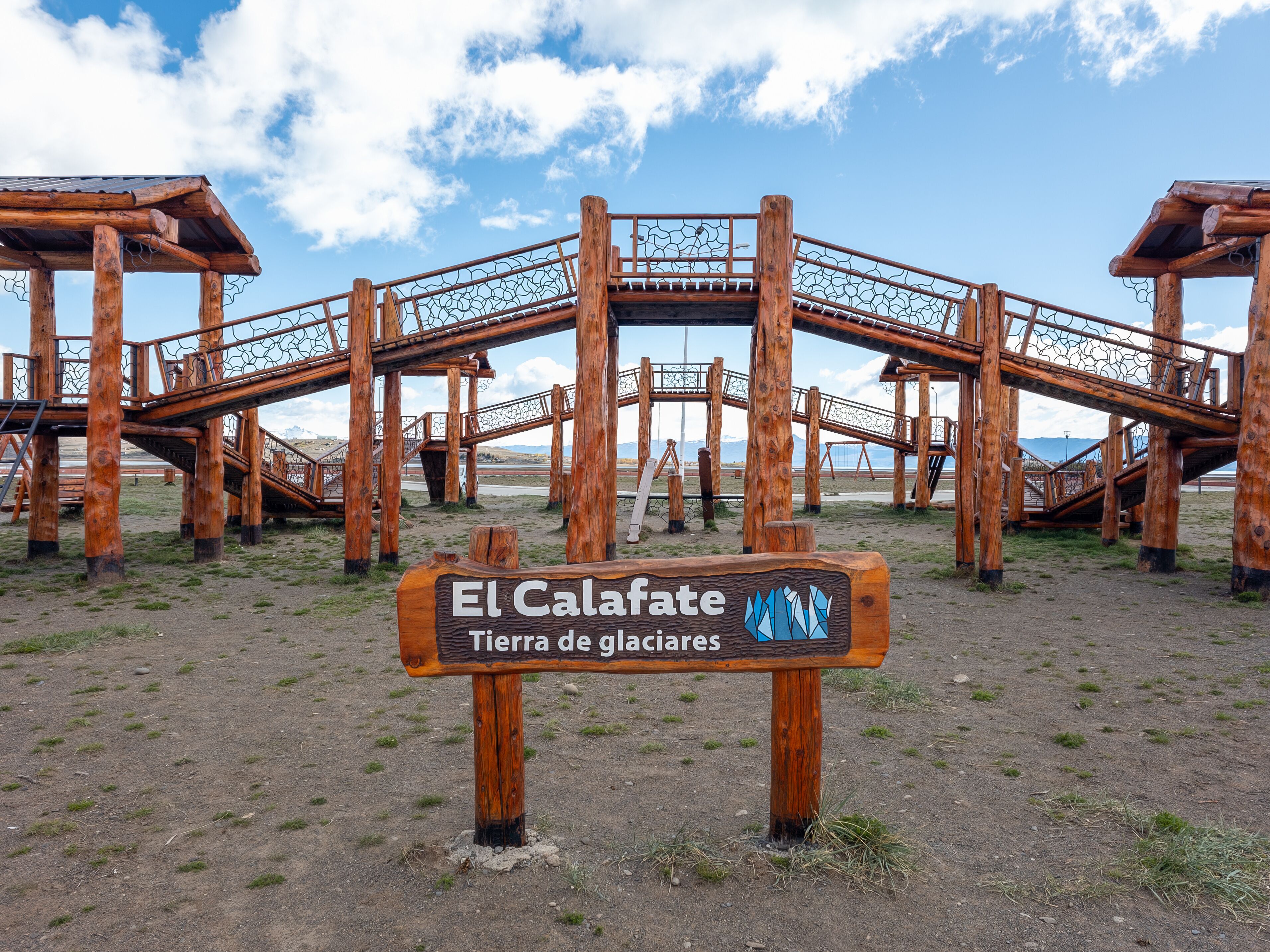 Uma placa de madeira com a inscrição 'El Calafate Tierra de Glaciares', rodeada por construções de madeira e um céu claro.
