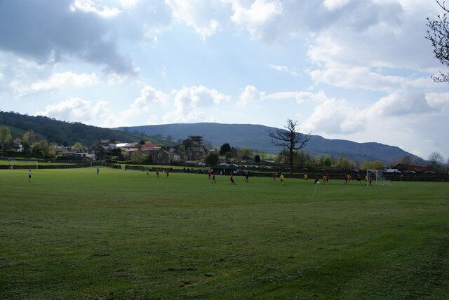 Um campo gramado com uma partida de futebol, casas e montanhas ao fundo.