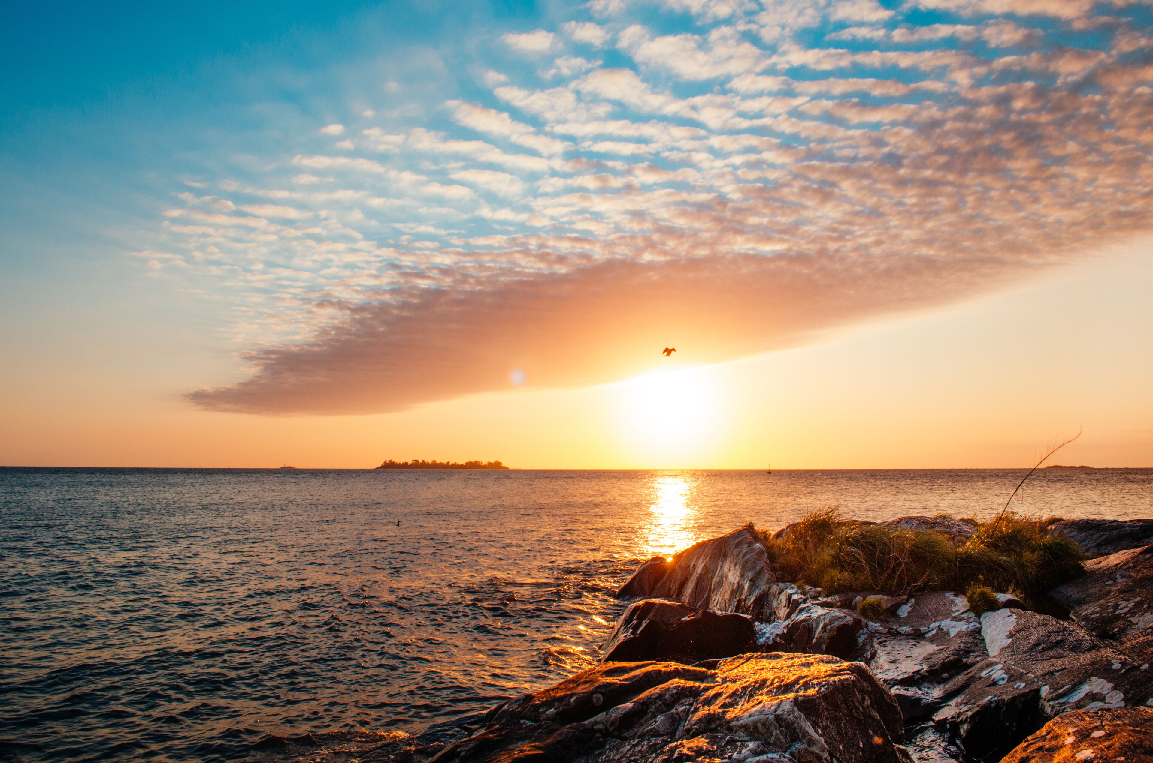 Pôr do sol sobre um mar calmo, com rochas em primeiro plano e uma ilha ao longe.