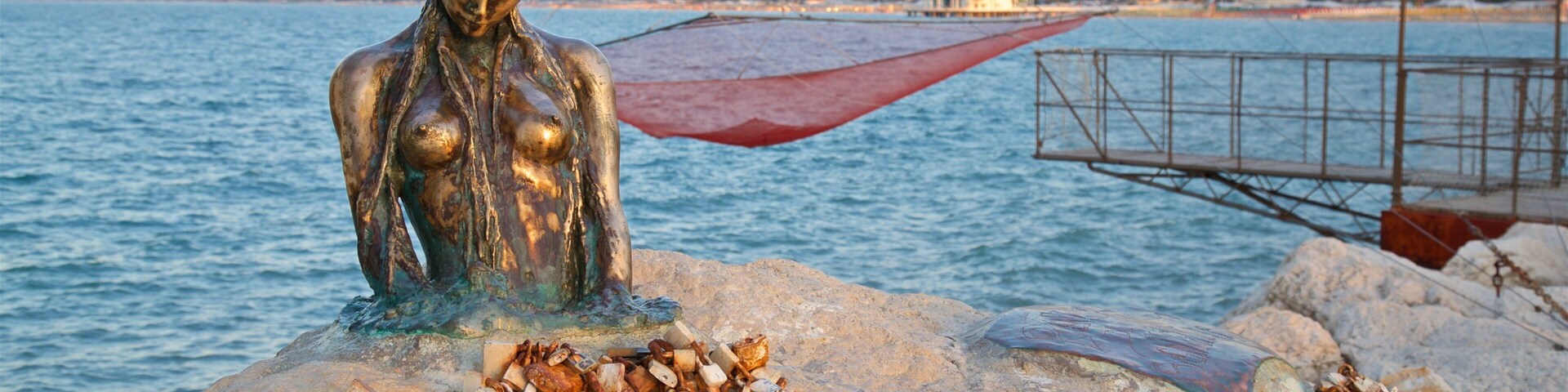 Estátua de uma mulher segurando um guarda-chuva vermelho em uma costa rochosa, com uma paisagem urbana ao fundo.