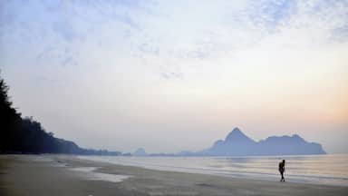 Uma praia com uma pessoa caminhando, águas calmas e montanhas ao longe.