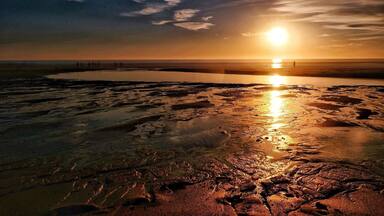 Um pôr do sol sobre uma praia molhada e refletora, com uma linha de costa distante e uma pessoa em frente à água.
