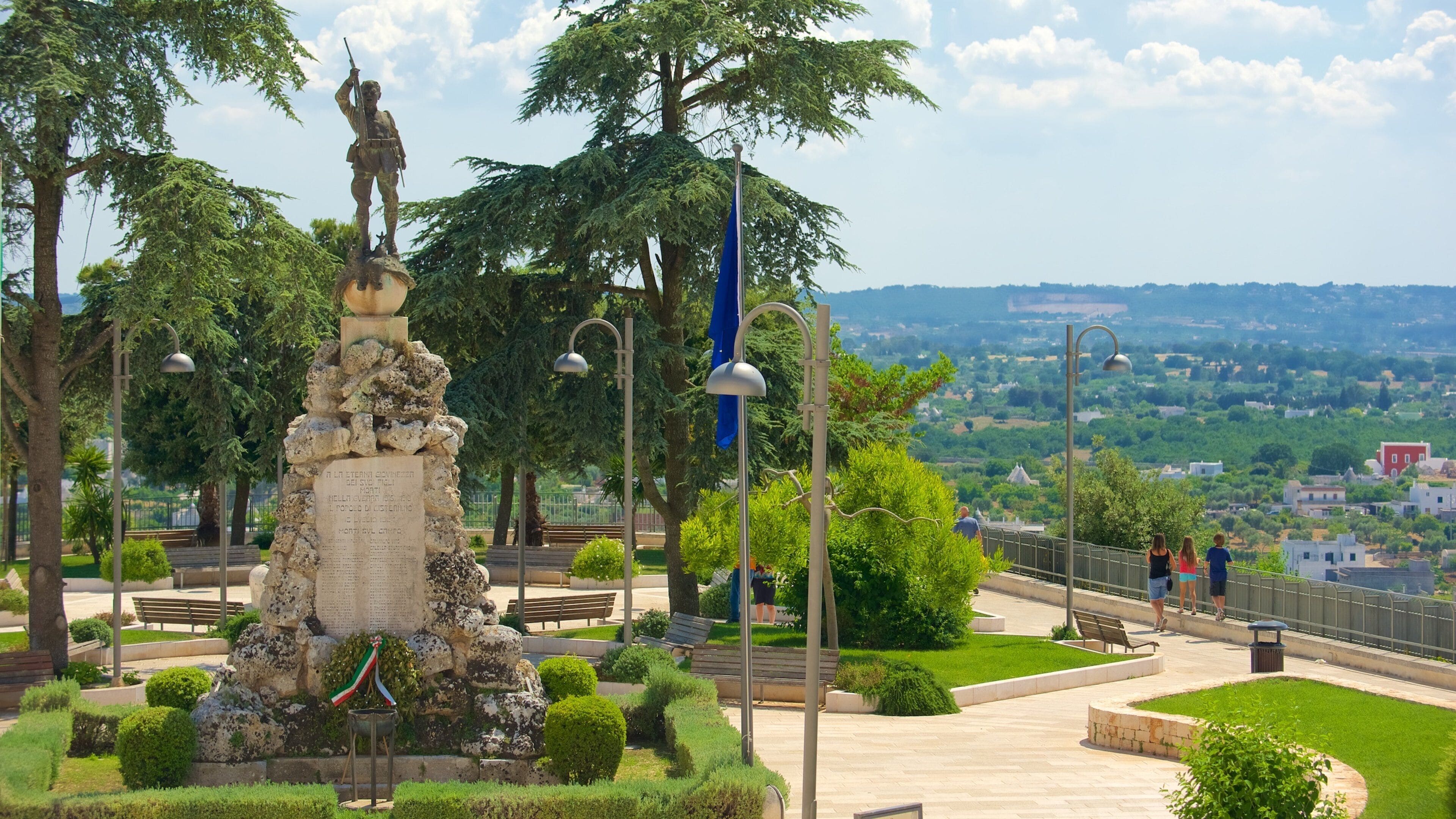 Um monumento com inscrições, cercado por vegetação bem cuidada e bancos, com vista para uma paisagem pitoresca e edifícios ao longe.