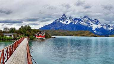 Um lago com uma ponte de madeira, uma casa vermelha e montanhas cobertas de neve ao fundo.