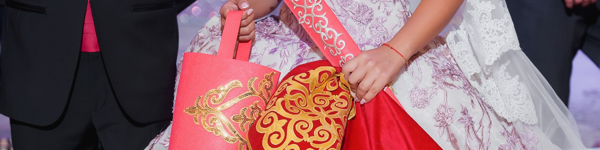 Uma pessoa num vestido branco com bordados vermelhos e dourados, segurando um leque cerimonial vermelho e dourado.