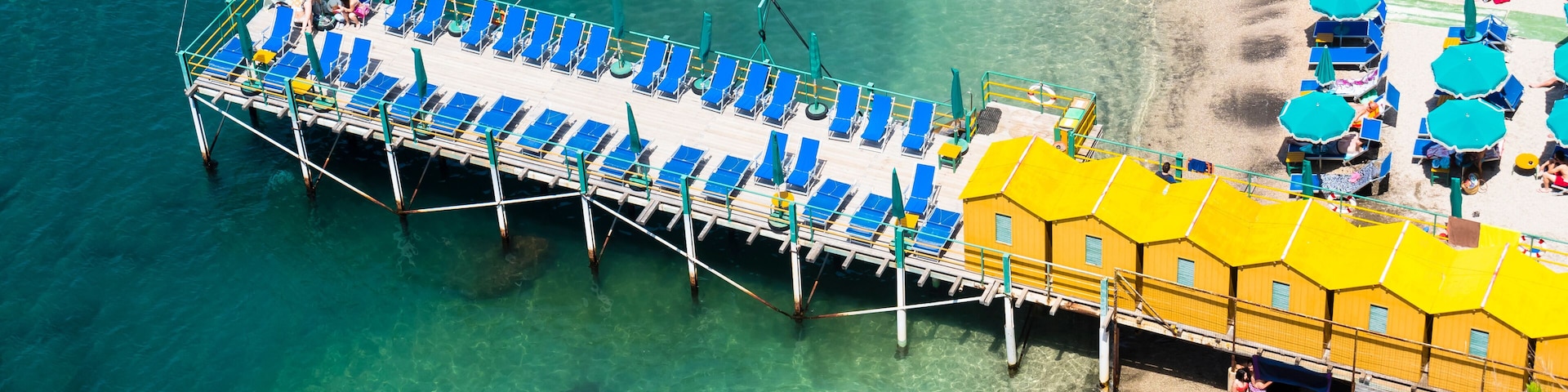 Um píer com cadeiras de banho azuis que se estende por águas turquesa cristalinas, uma praia arenosa com guarda-sóis e um edifício amarelo.