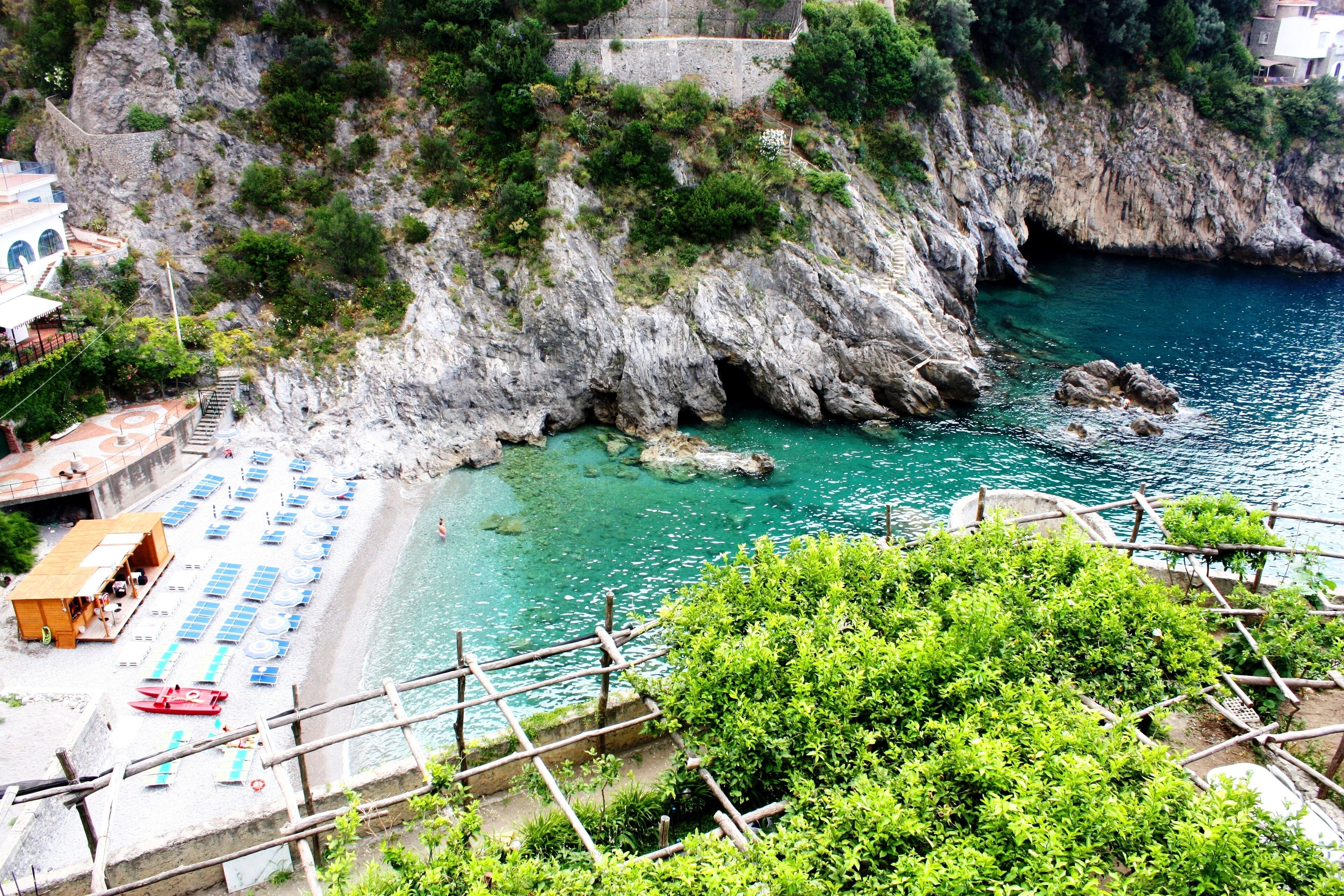 Uma área costeira com um penhasco rochoso, águas turquesa cristalinas e uma praia com guarda-sóis e uma pequena estrutura de madeira.