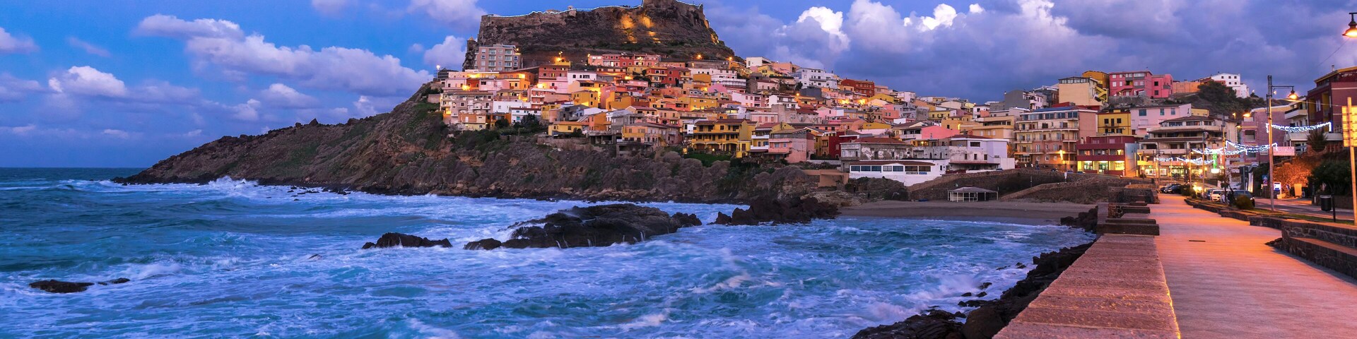 Uma cidade costeira com edifícios coloridos, uma fortaleza imponente numa colina e uma linha costeira rochosa.