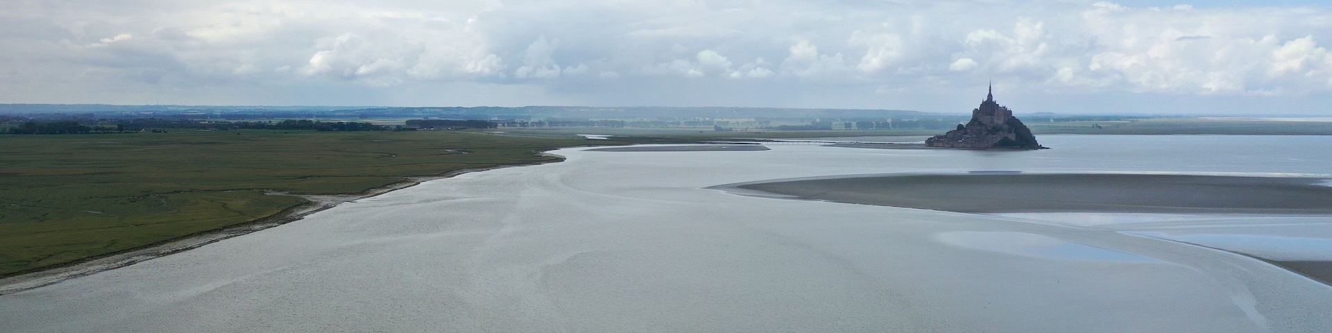 Paisagem costeira com uma grande massa de água, uma pequena ilha com uma construção semelhante a um castelo e colinas distantes sob um céu nublado.