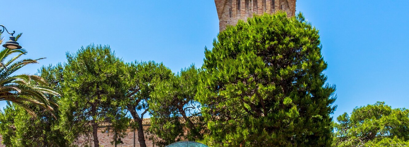 Uma torre histórica com uma bandeira, cercada por vegetação exuberante e uma grande escultura esférica.