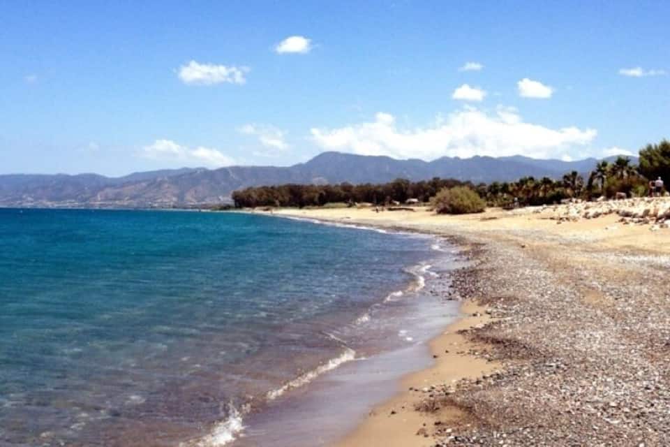 Uma praia com águas cristalinas, areal de pedras e montanhas ao longe.