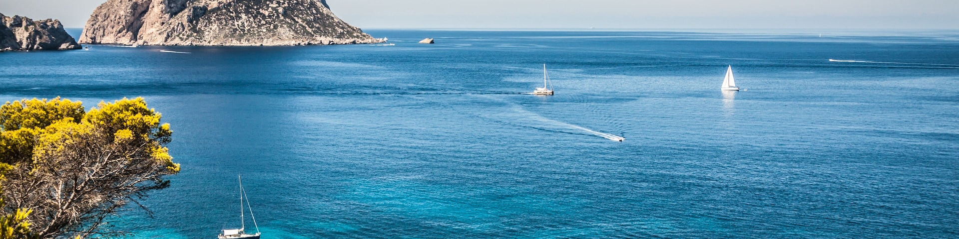 Mar azul cristalino com barcos, uma ilha rochosa e vegetação exuberante.