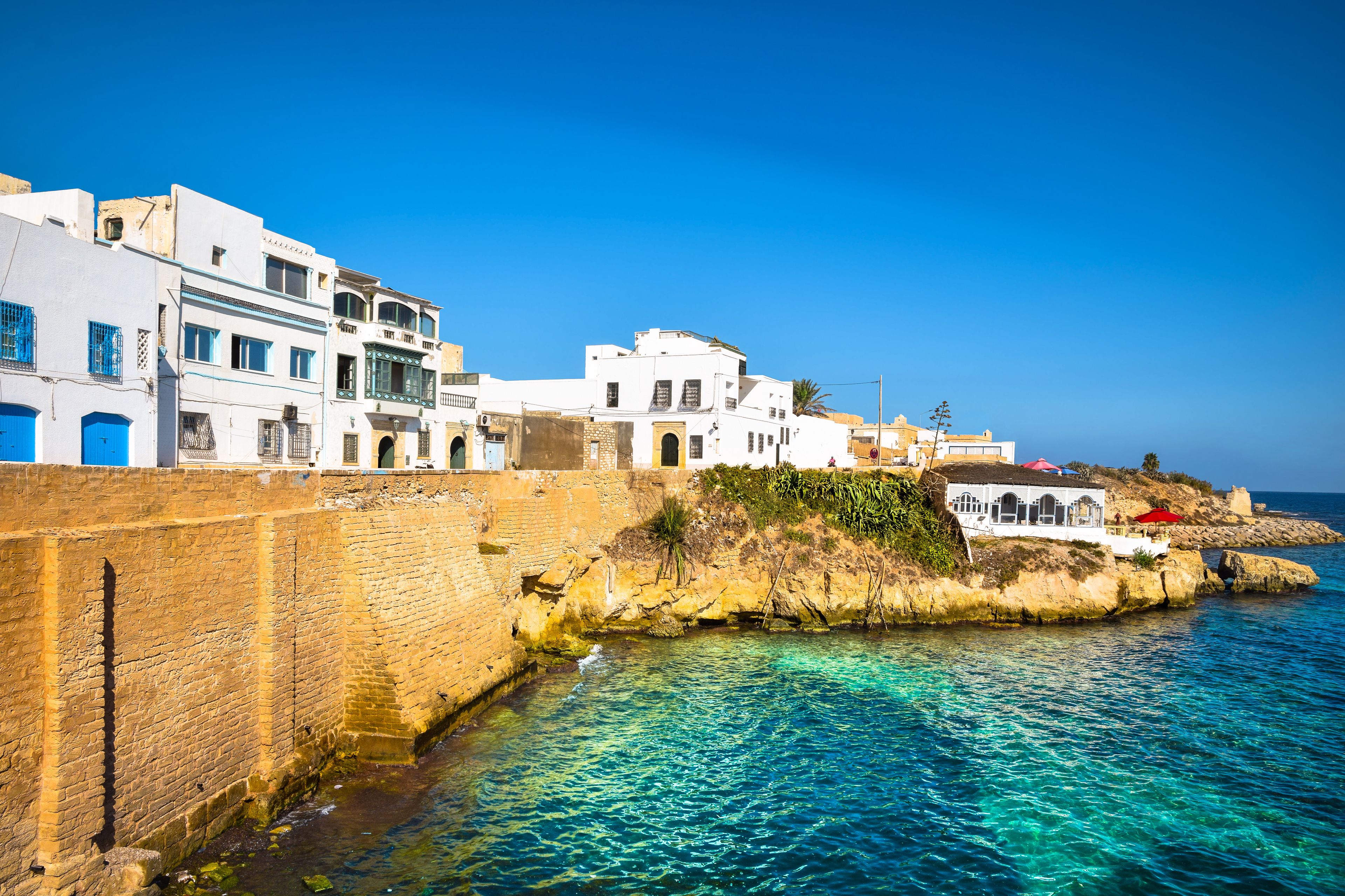 Uma cidade costeira com edifícios brancos, águas azul-claras e uma orla rochosa.