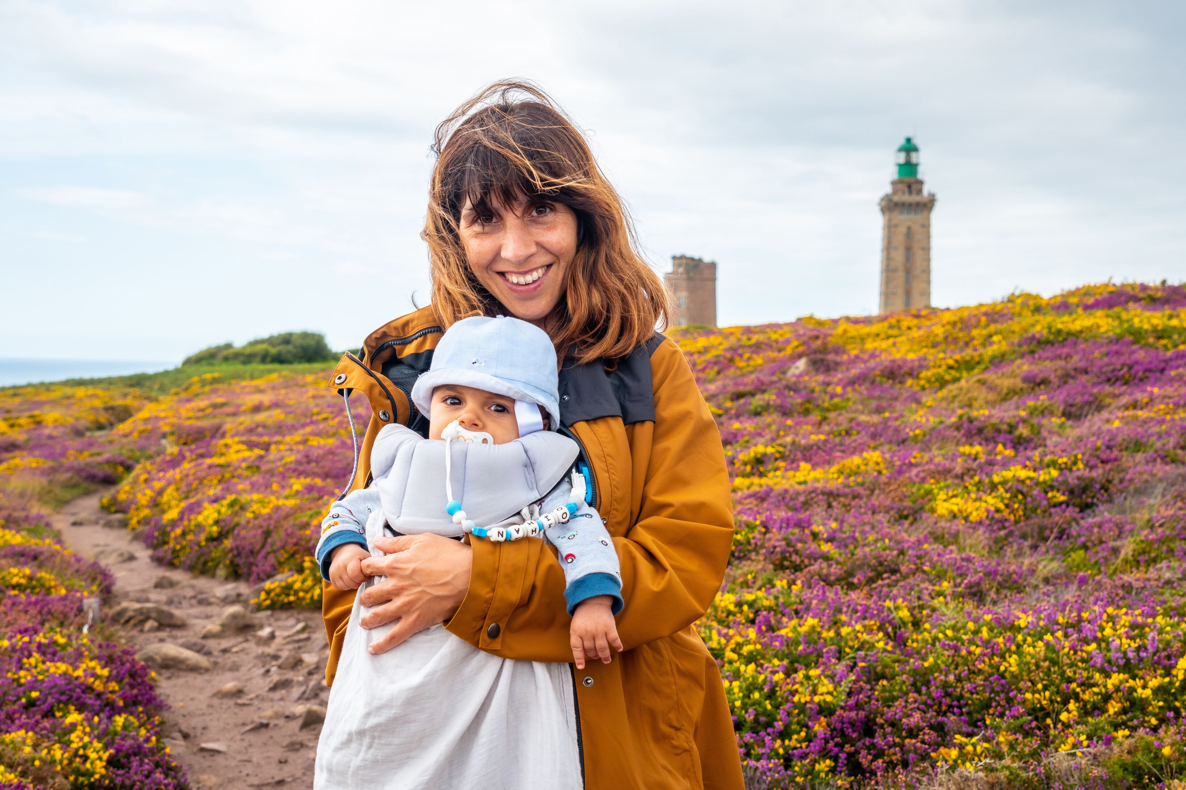 Uma pessoa segura um filho em um campo de flores roxas e amarelas, com um farol ao fundo.