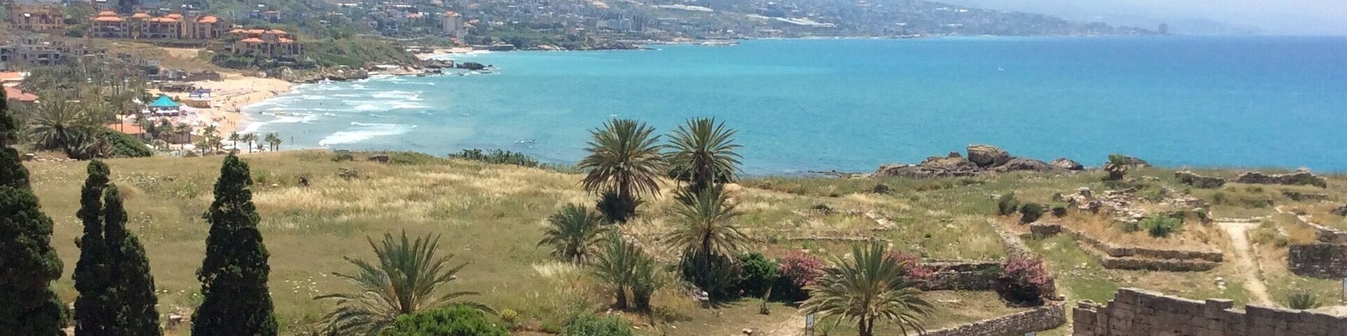 Paisagem costeira com praia, mar azul límpido e uma cidade montanhosa ao fundo.