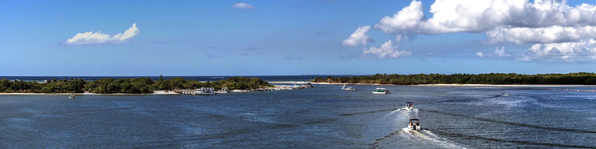 Cena costeira com céu claro, um barco deixando rastro e um litoral distante com árvores e edifícios.