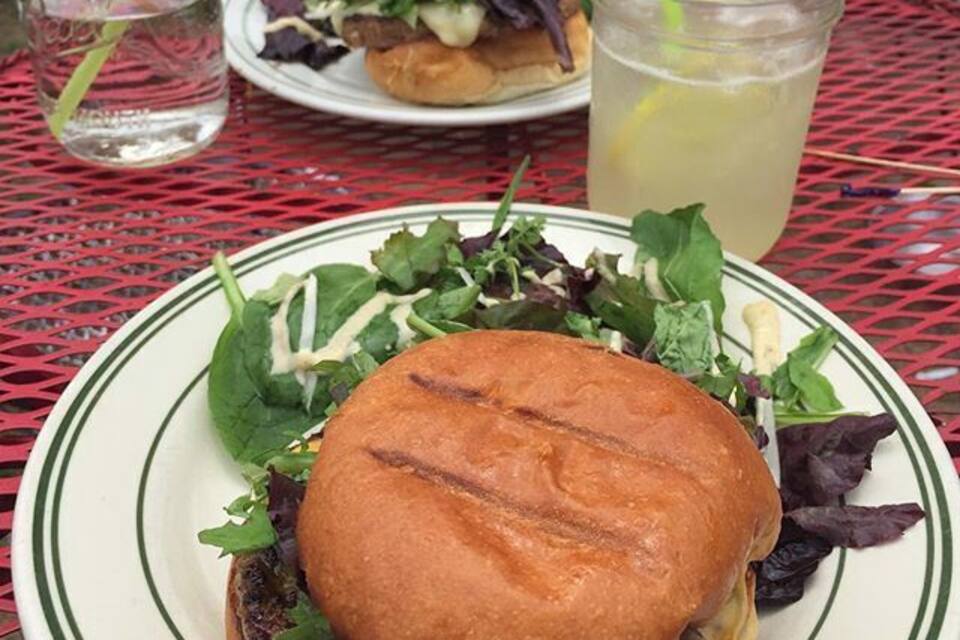 Um hambúrguer com alface e queijo em um prato, um copo de limonada e uma bebida com canudo sobre uma mesa de metal vermelha.