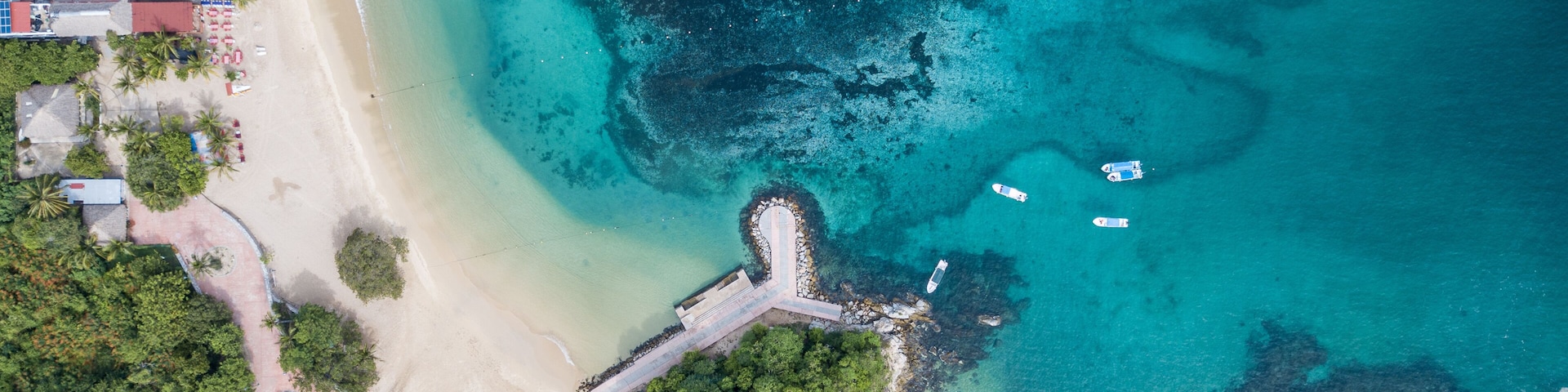 Vista aérea de uma área costeira com uma praia arenosa, águas turquesa cristalinas e um pequeno cais.