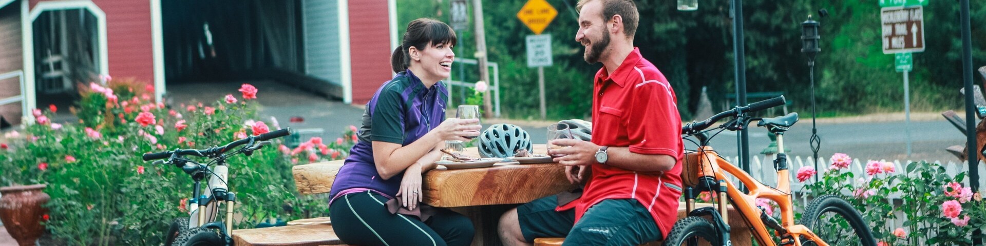 Duas pessoas sentadas em uma mesa de madeira ao ar livre, com bicicletas por perto, cercadas por flores e vegetação.