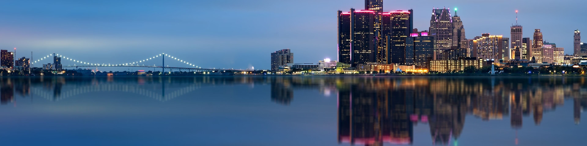 Linha do horizonte da cidade com edifícios iluminados e uma ponte refletida na água.