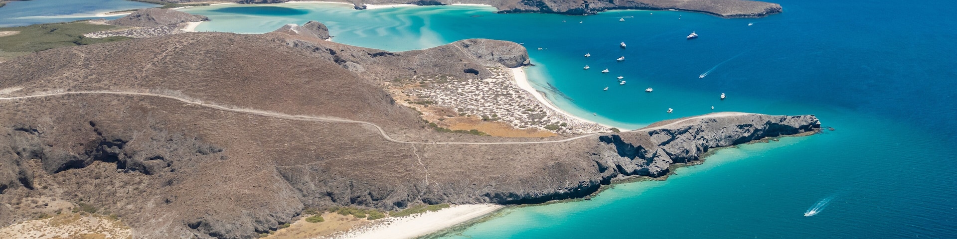 Paisagem costeira com praia, águas turquesa cristalinas e montanhas ao fundo.
