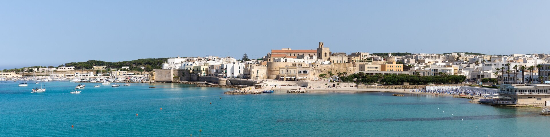Uma vila costeira com um castelo histórico, barcos e um mar azul límpido.