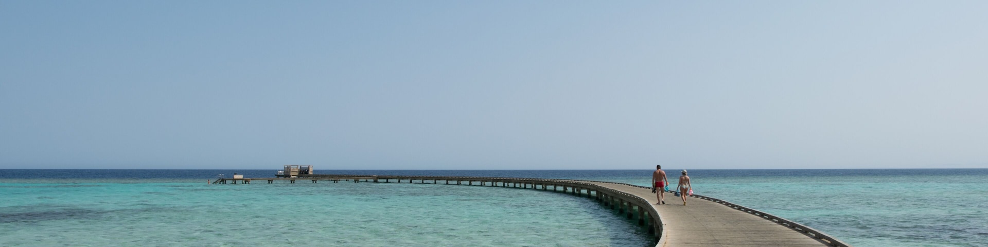 Um cais de madeira que se estende por águas turquesa e cristalinas, com algumas pessoas caminhando.
