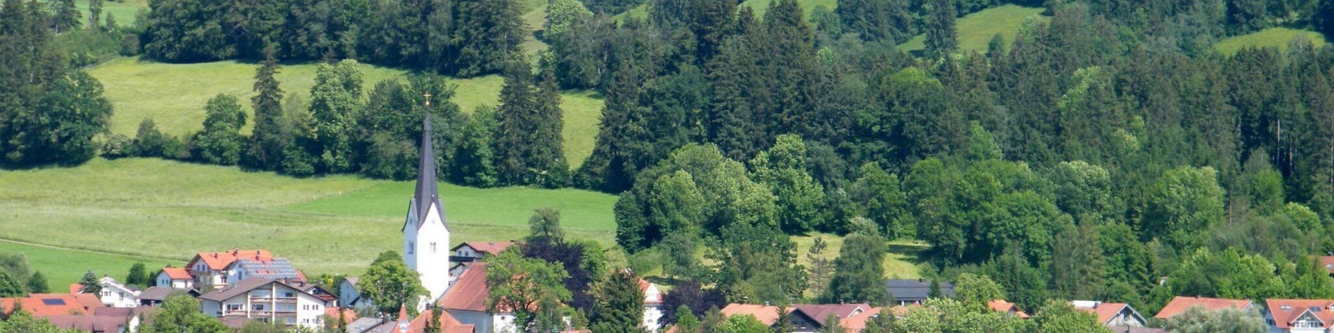 Paisagem rural com colinas ondulantes, uma igreja com uma torre alta e edifícios residenciais com telhados vermelhos.
