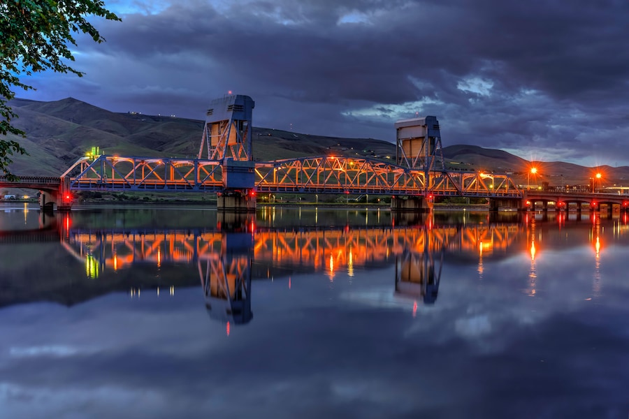 Uma ponte com estrutura treliçada, refletida em águas calmas, com colinas ao fundo.