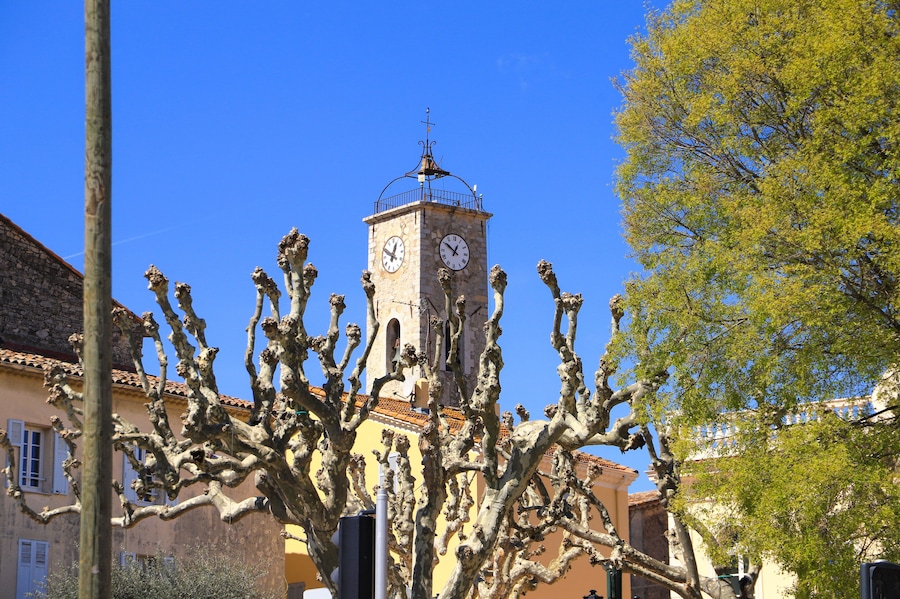 Uma torre com um relógio, rodeada por edifícios e uma árvore sem folhas.