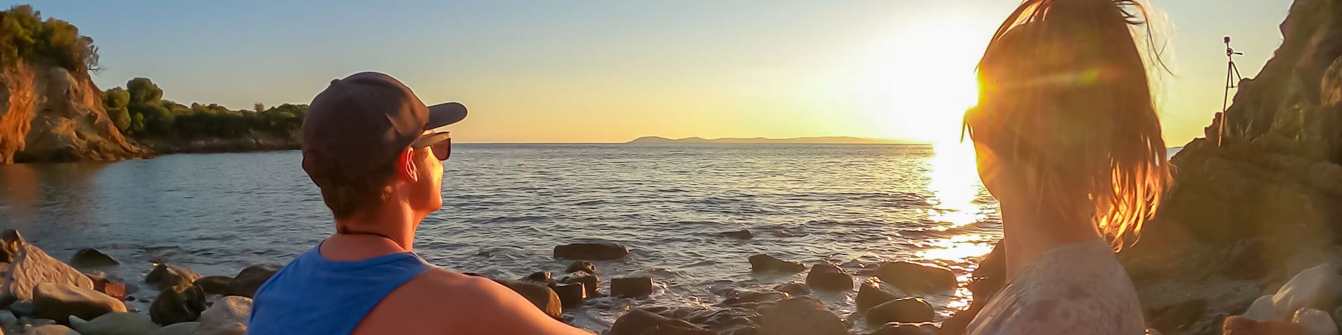 Um casal sentado numa praia rochosa ao pôr do sol, com o oceano ao fundo.