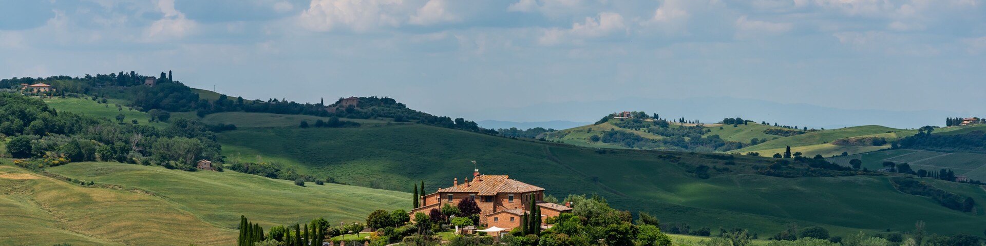 Colinas verdes e ondulantes, uma casa rústica rodeada de árvores, e colinas distantes sob um céu parcialmente nublado.