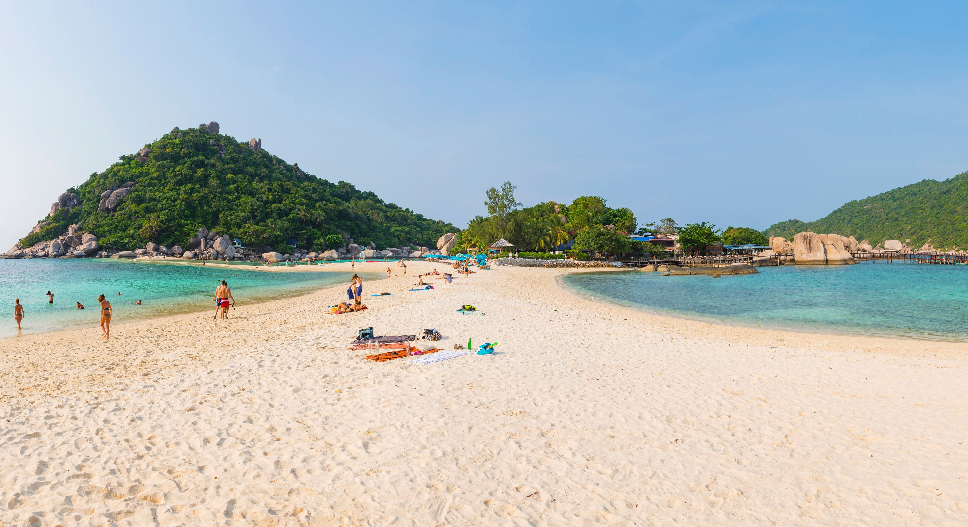 Uma praia com águas turquesa cristalinas, areal arenoso e uma grande formação rochosa ao fundo.