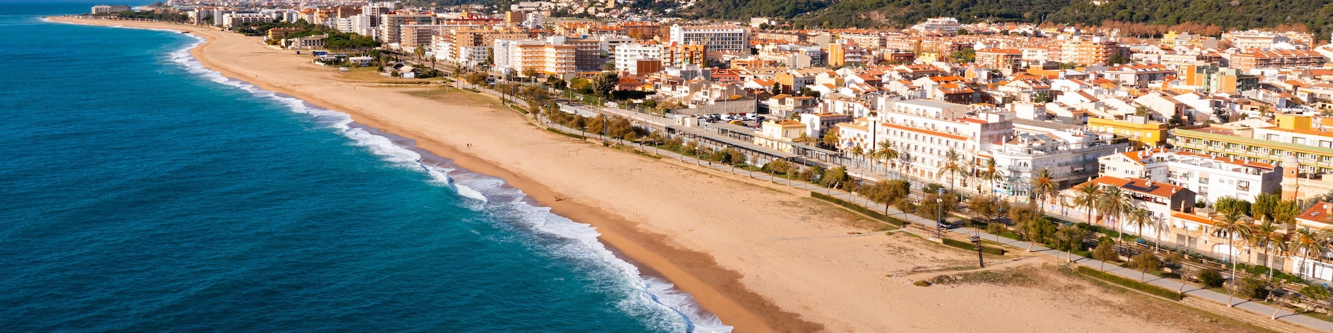Uma cidade costeira com uma extensa praia de areia, área urbana densa e colinas verdes ao fundo.