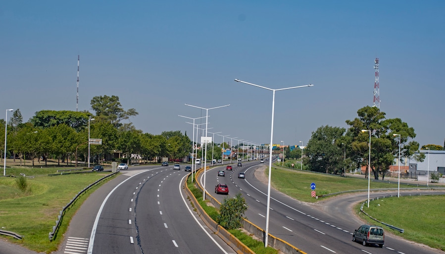 Uma autoestrada com múltiplas faixas, iluminação pública, uma curva e vegetação nas laterais.